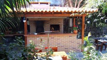 Área de churrasco de tijolos ao ar livre com pérgola de madeira, rodeada por vasos de plantas e uma mesa de jantar.