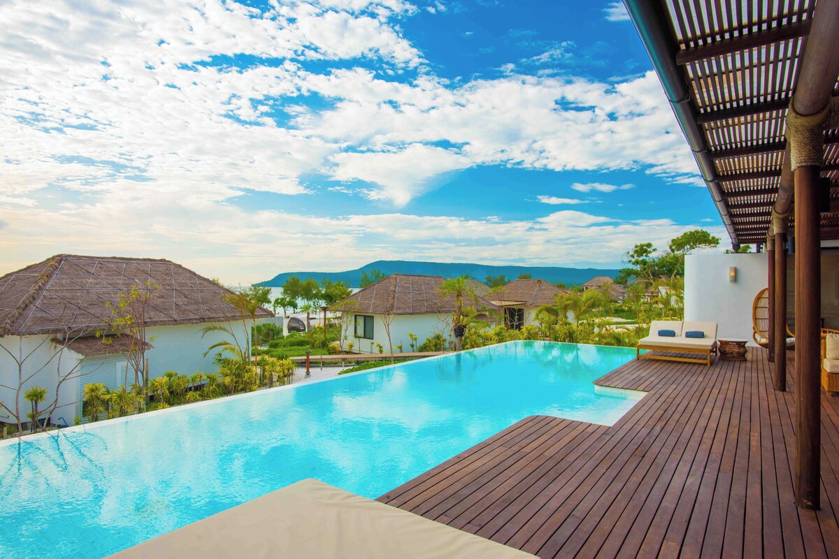 Área de piscina com deck de madeira, espreguiçadeiras e estruturas com telhado de palha.
