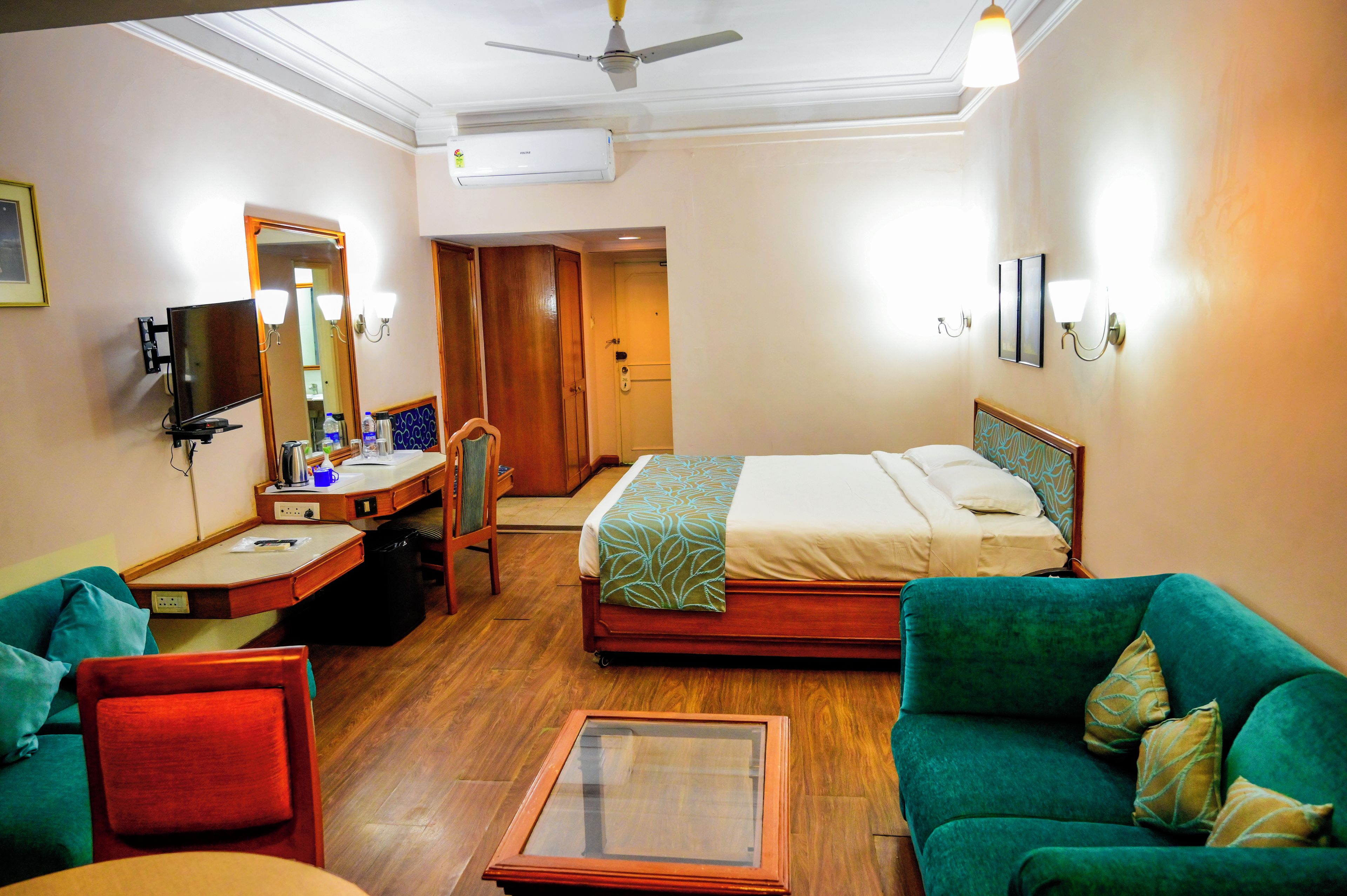 Quarto de hotel com cama, escrivaninha, cadeira, televisão e ventilador de teto.