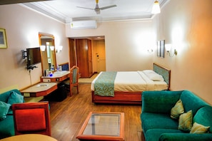 Quarto de hotel com cama, escrivaninha, cadeira, televisão e ventilador de teto.
