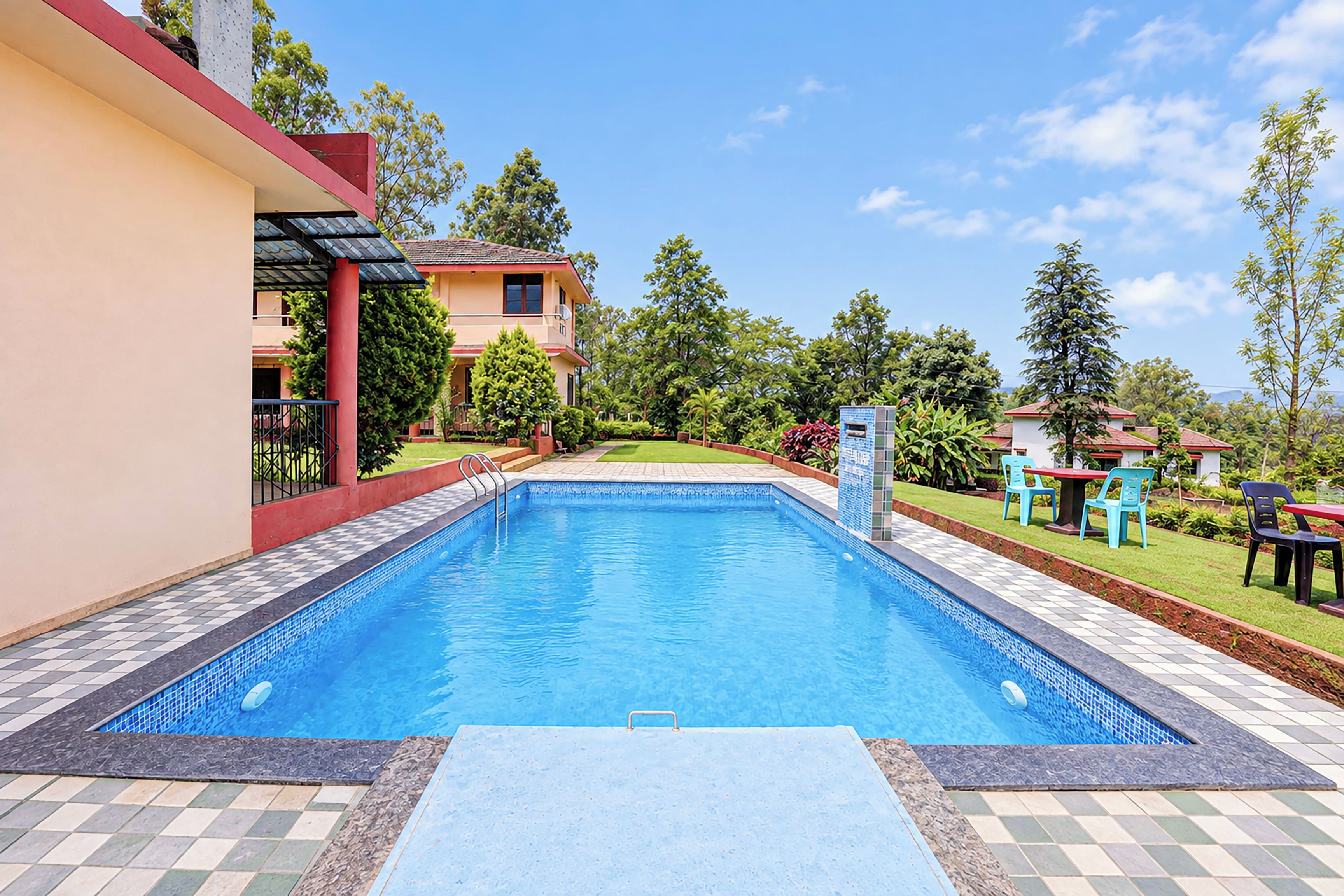 Piscina com deck revestido, rodeada por vegetação e um edifício com telhado vermelho.