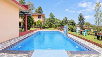 Piscina com deck revestido, rodeada por vegetação e um edifício com telhado vermelho.