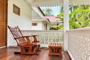 Uma cadeira de balanço de madeira e um banquinho em um alpendre com guarda-corrimão branco, com vista para uma casa com telhado vermelho e vegetação exuberante.