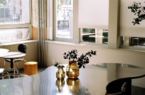 Interior moderno com mesa de vidro, dois vasos dourados com plantas, uma cadeira e janela com vista para edifícios.