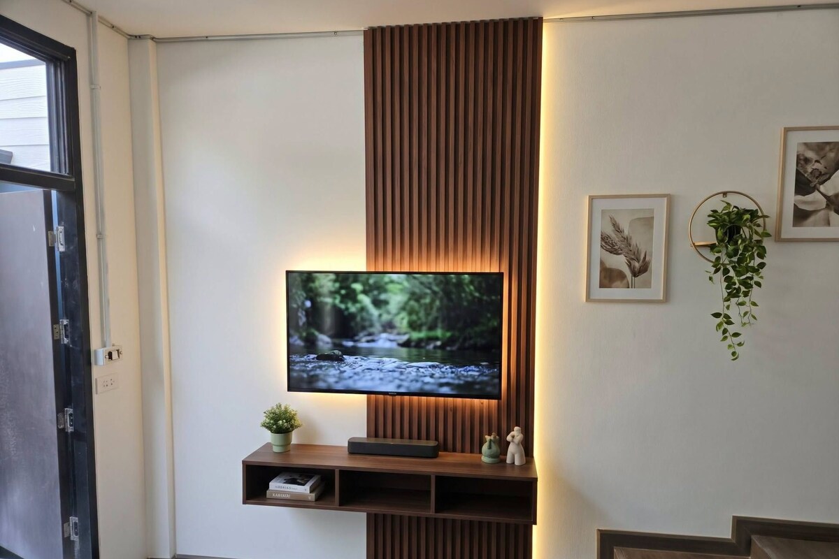 Televisão de tela plana presa à parede, com painel de madeira, uma pequena prateleira com objetos decorativos e um quadro à direita.