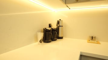 Quarto de hotel com bancada branca, cafeteira, chaleira e uma pilha de copos.