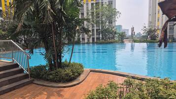Piscina com deck de madeira e guarda-corpo de metal, rodeada por vegetação e edifícios modernos ao fundo.