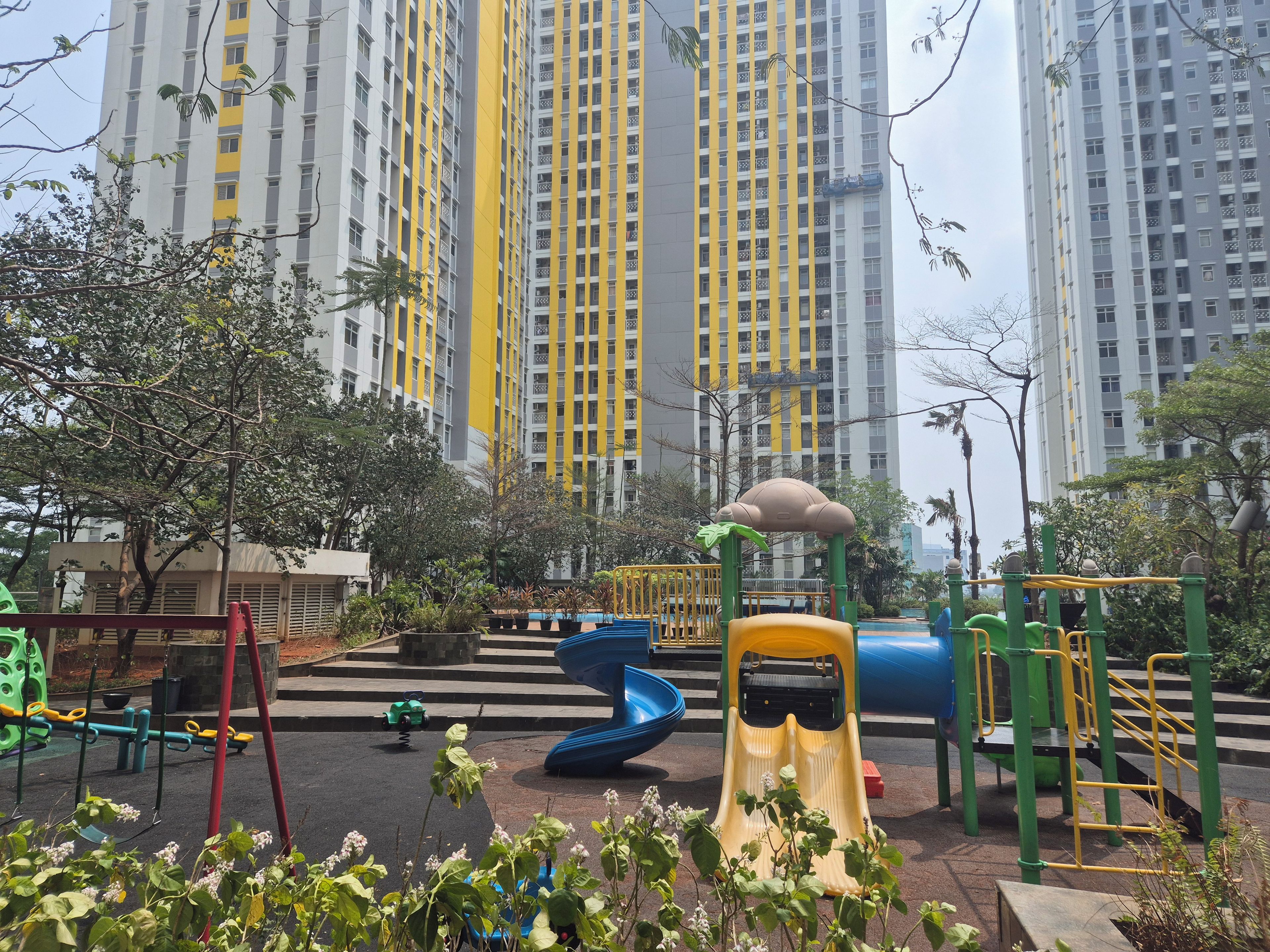 Um parquinho com diversos equipamentos de recreação, cercado por árvores e edifícios ao fundo.