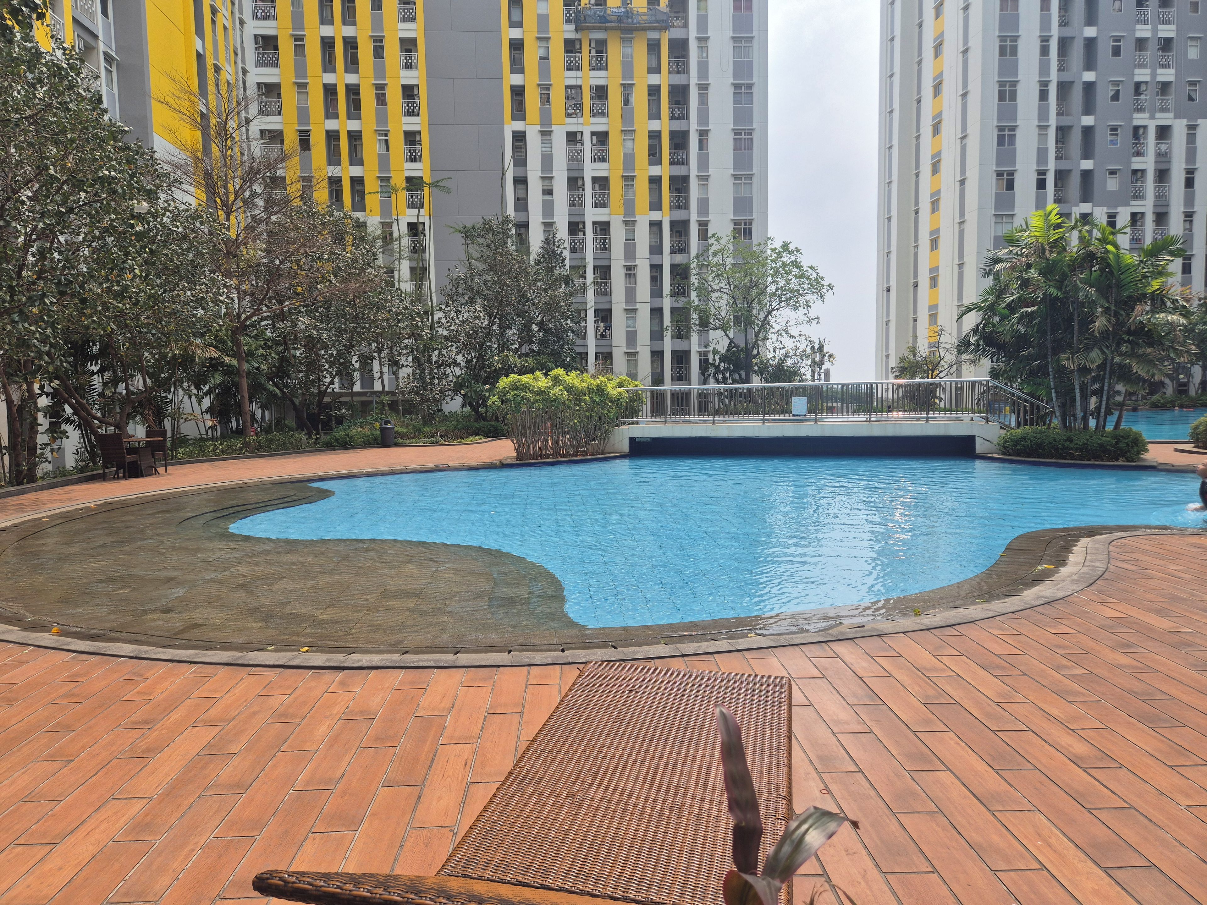 Piscina com deck de tijolos, cercada por vegetação e edifícios residenciais altos.