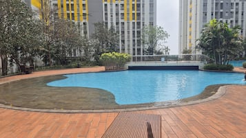Piscina com deck de tijolos, cercada por vegetação e edifícios residenciais altos.