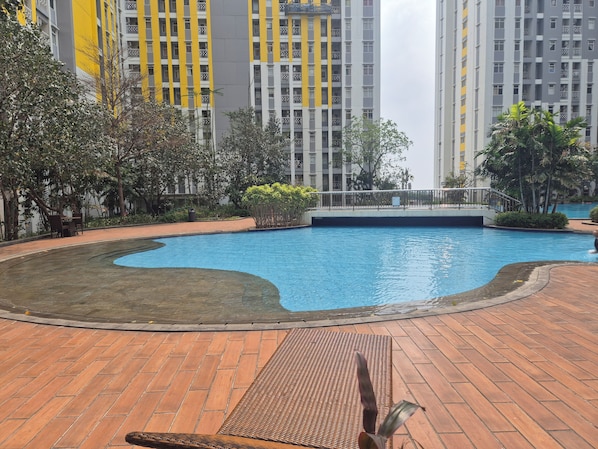 Piscina com deck de tijolos, cercada por vegetação e edifícios residenciais altos.