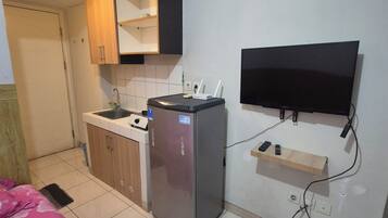 Uma cozinha compacta com geladeira, micro-ondas e pia. Há uma televisão fixada na parede e uma pequena prateleira abaixo.