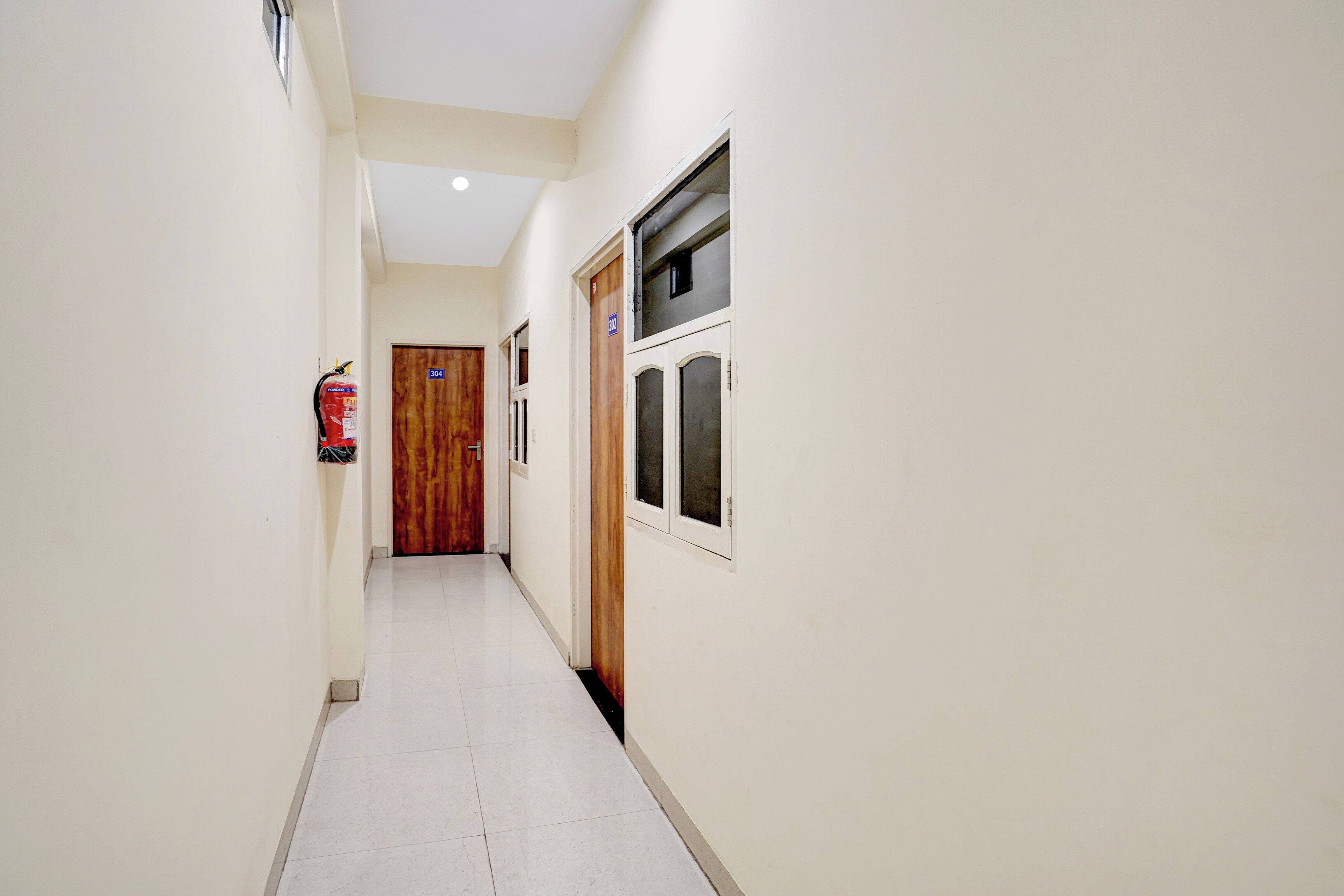 Um corredor com um extintor fixado na parede, uma porta de madeira com placa de identificação e uma janela com moldura de metal.