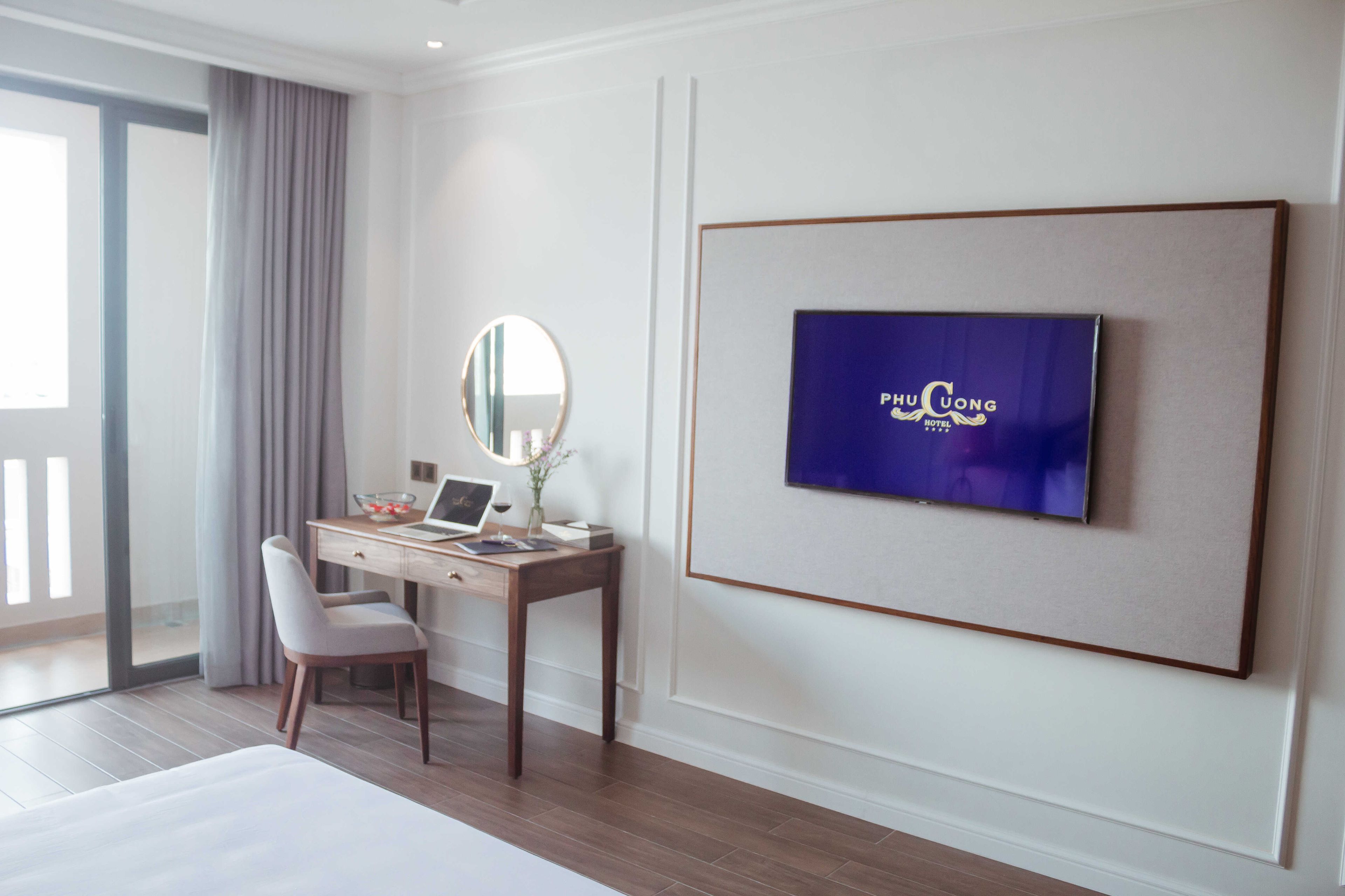Quarto de hotel com uma cama, uma escrivaninha com um laptop, um espelho e uma televisão na parede.