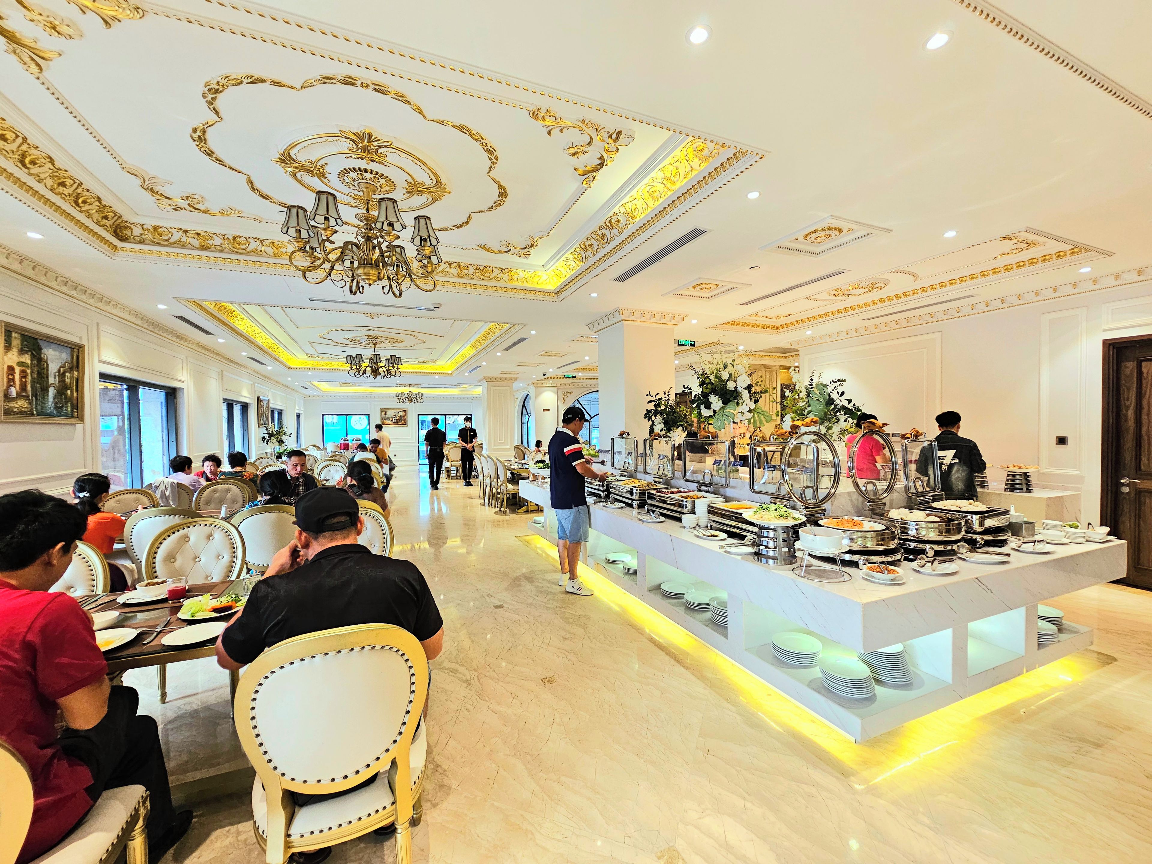 Espaço de refeições de hotel com decoração de teto elaborada, balcão de buffet e hóspedes a jantar.