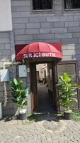 Entrada de um prédio com marquise vermelha identificada como 'SUR ICI BUTIK' e uma tampa de bueiro no caminho de pedras.