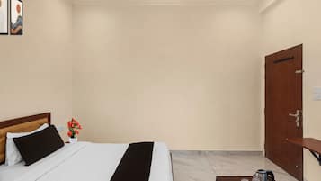 Quarto de hotel com cama, mesa de cabeceira, um vaso com flores, porta de madeira e uma pequena mesa com chaleira e xícaras.