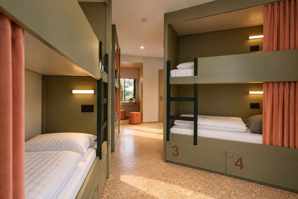 Quarto de hotel com beliches, janela com cortinas, uma mesinha e luminária na parede.