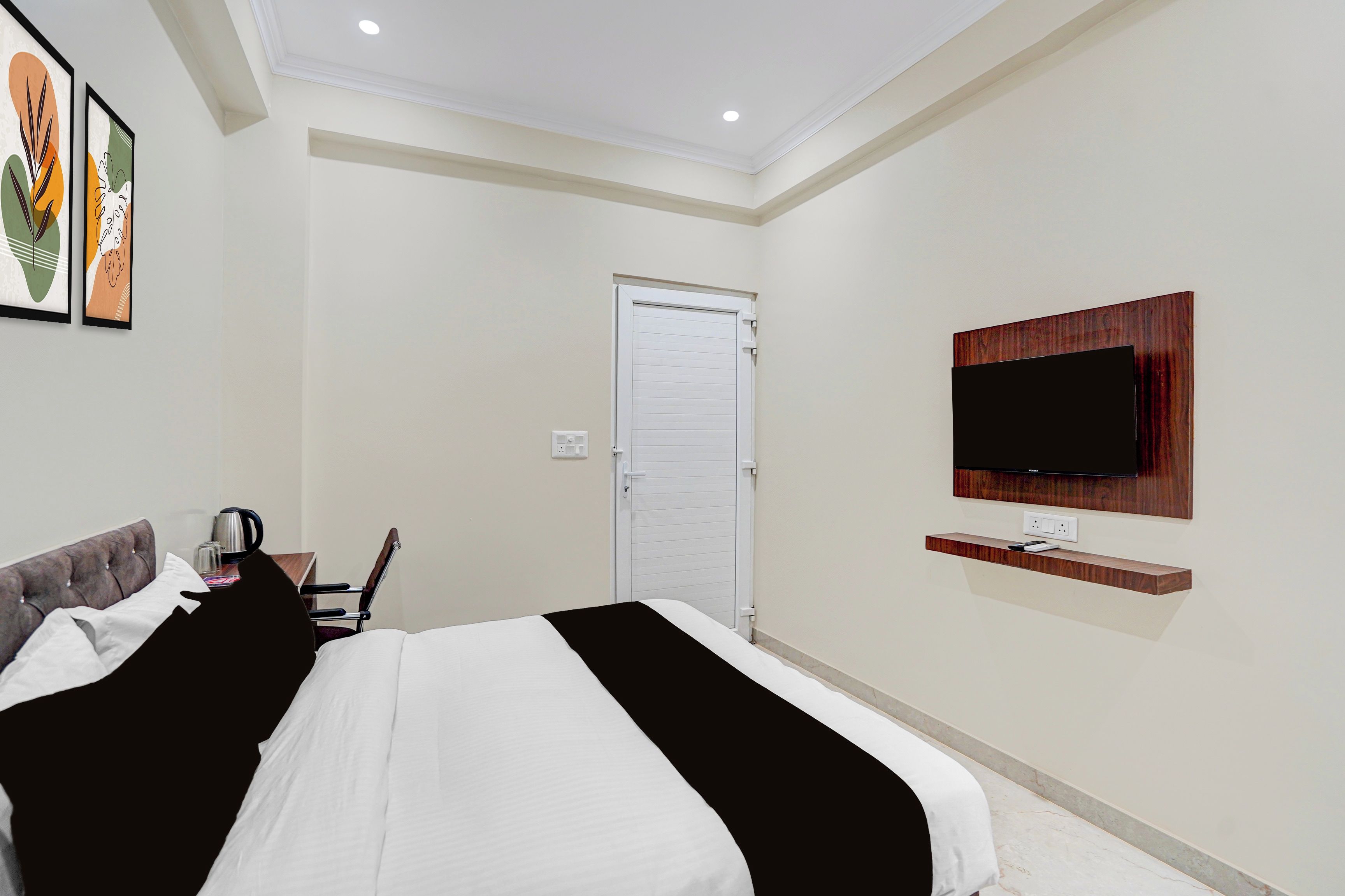 Quarto de hotel com uma cama, televisão presa na parede, uma porta e uma pequena prateleira com controle remoto.