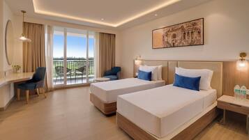 Quarto de hotel com duas camas, uma escrivaninha, uma cadeira, varanda com vista e um quadro na parede.