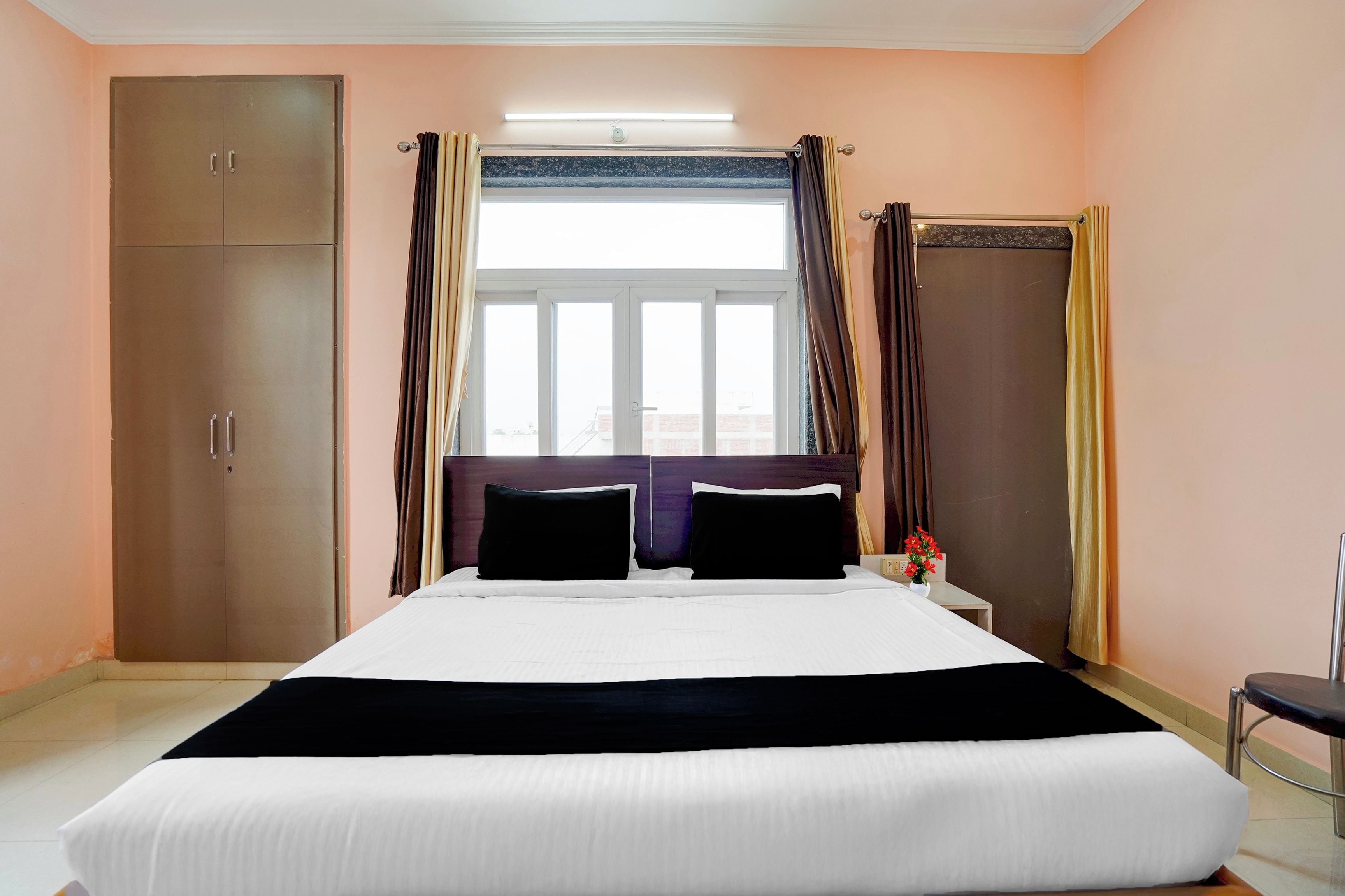 Quarto de hotel com uma cama grande, um guarda-roupa, uma janela com cortinas e uma mesa de cabeceira com um vaso de flores.