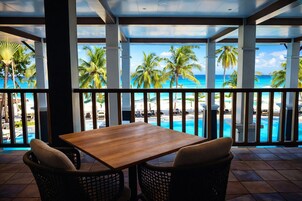 Mesa de madeira com duas cadeiras, vista para área da piscina com palmeiras e praia, e céu claro.
