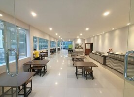 Ampla área de refeições de hotel, com várias mesas e cadeiras, um balcão de recepção e grandes janelas que proporcionam luz natural.