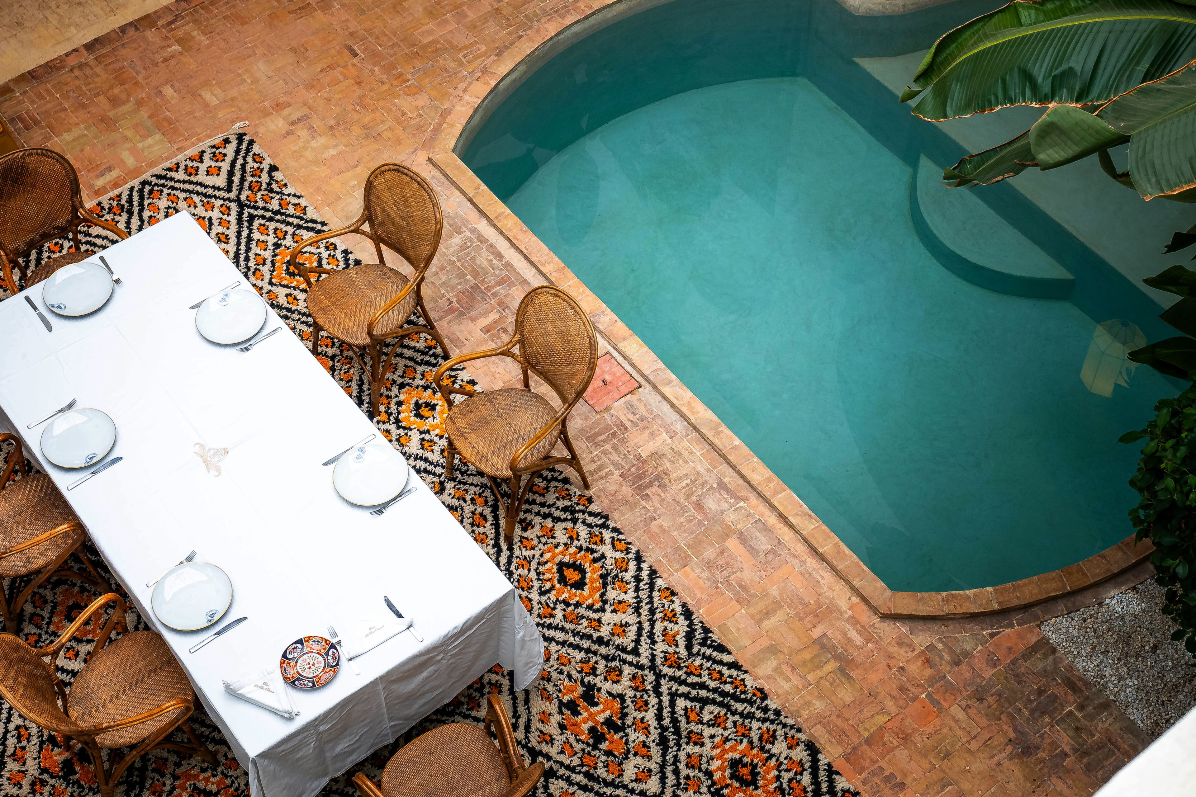 Área de refeições à beira da piscina com mesa posta para seis, rodeada por cadeiras de vime.