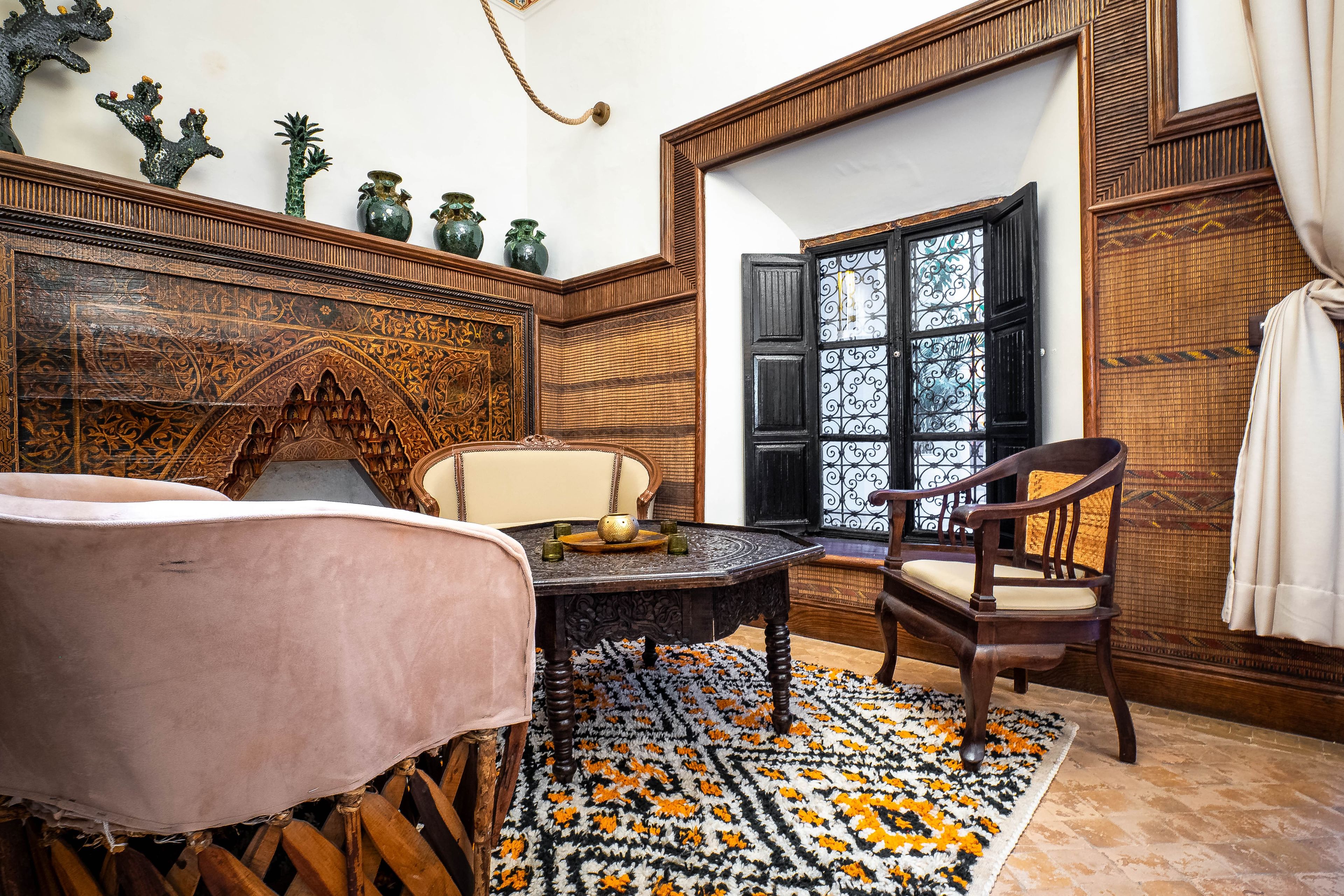 Quarto tradicional com cabeceira de madeira entalhada, uma mesa redonda e um tapete estampado, além de uma janela com grades de ferro decorativas.