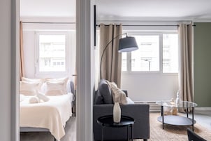 Quarto de hotel com cama, sofá, mesinha, abajur e vista para a cidade pela janela.