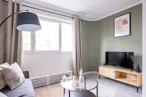Uma sala de estar moderna com sofá, mesa de centro de vidro, TV sobre suporte de madeira e uma grande janela com cortinas.