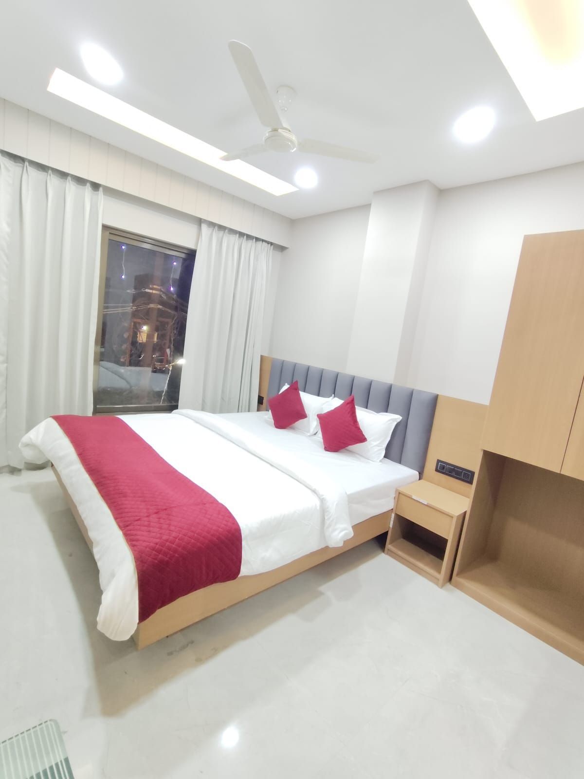 Quarto de hotel com cama, mesa de cabeceira, ventoinha no teto e janela com cortinas.