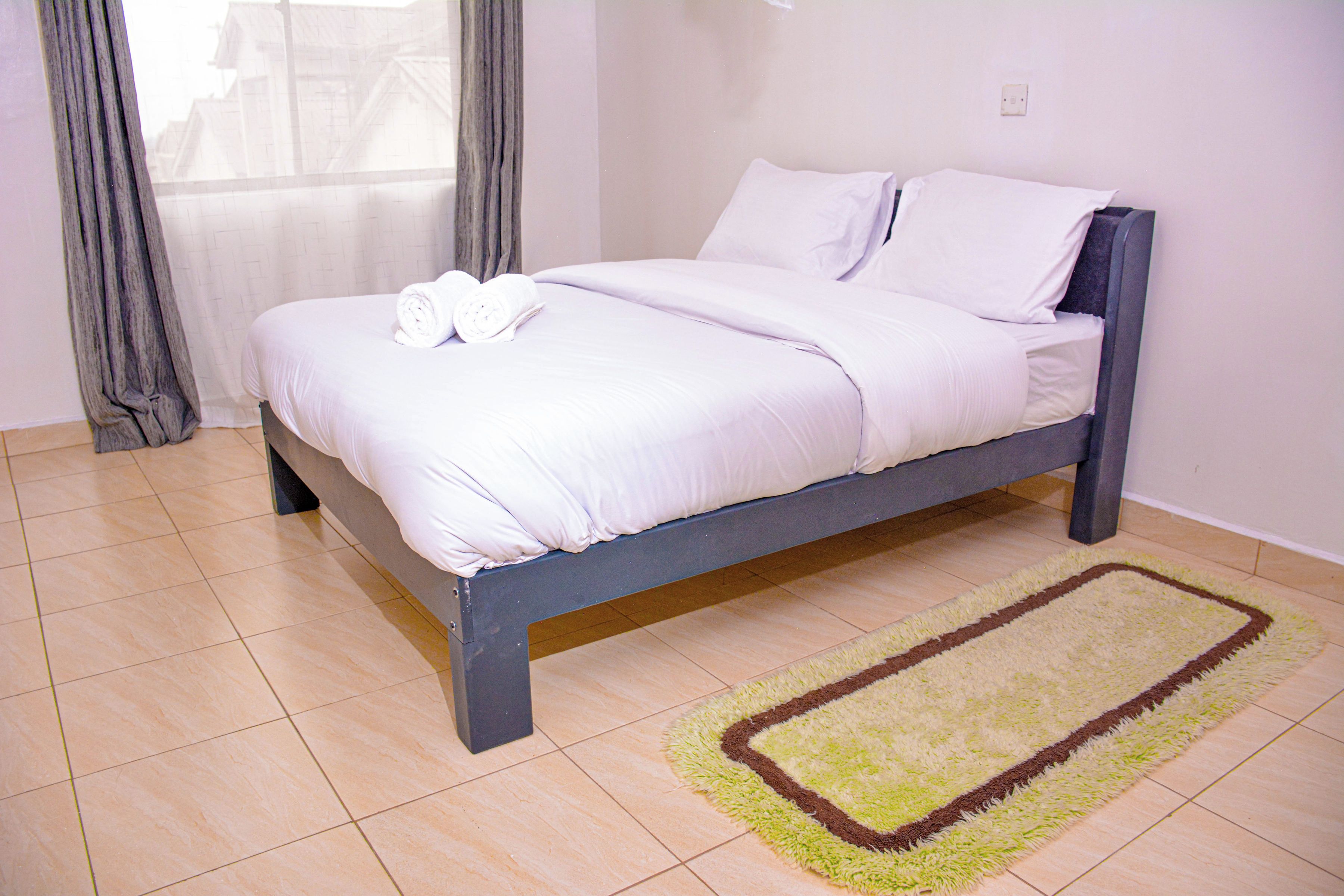 Cama com lençóis brancos e toalha enrolada, cabeceira cinzenta e tapete claro sobre piso de azulejos.