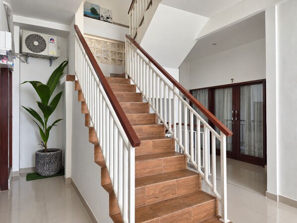 Uma escadaria com degraus de madeira e guarda-corpos brancos, conduzindo a um patamar com um painel decorativo e um ventilador de teto.