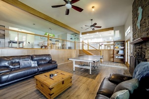 Uma sala de estar ampla com sofá de couro, mesa de centro de madeira e mesa de pingue-pongue. Uma escadaria dá acesso a um andar superior com área de cozinha.
