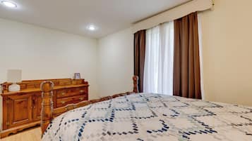 Quarto com cômoda de madeira, cama com colcha estampada e janela com cortinas.