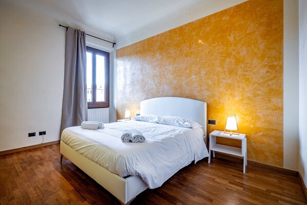 Quarto com cama, mesa de cabeceira, luminária e janela com cortinas.