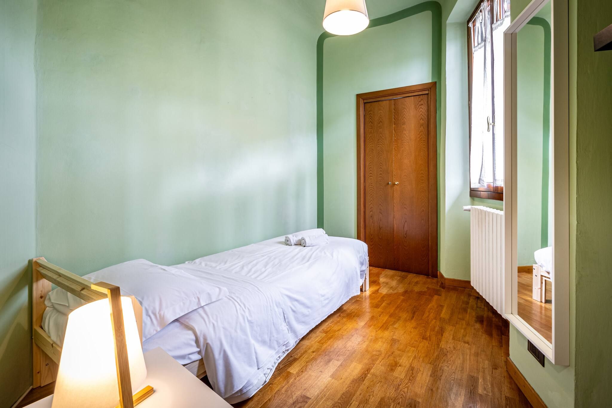 Quarto pequeno e arrumado com uma cama de solteiro, porta de madeira e janela com vista.