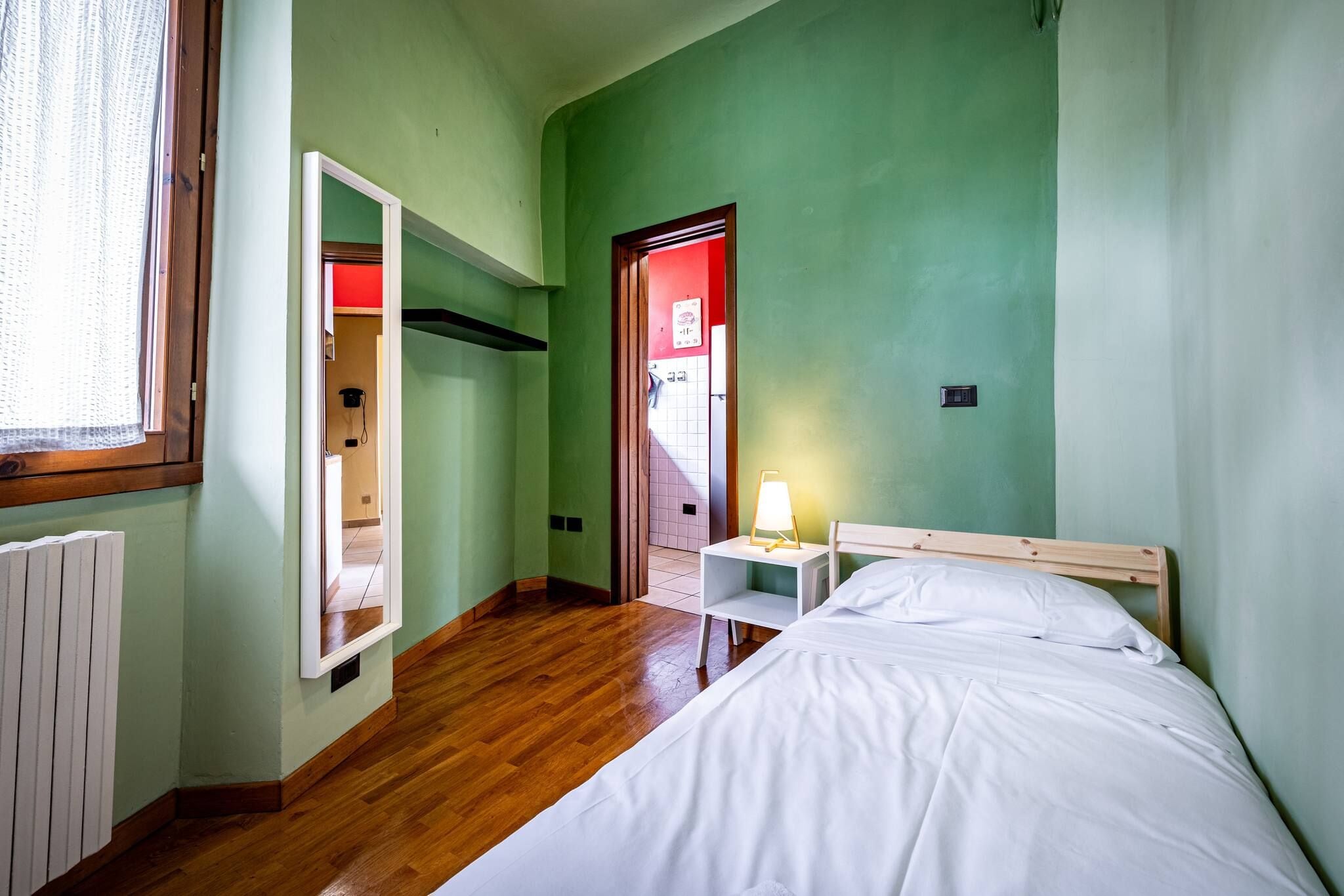 Um quarto com piso de madeira, uma cama com roupa de cama branca, uma mesa de cabeceira com abajur e uma porta que dá para outro cômodo.