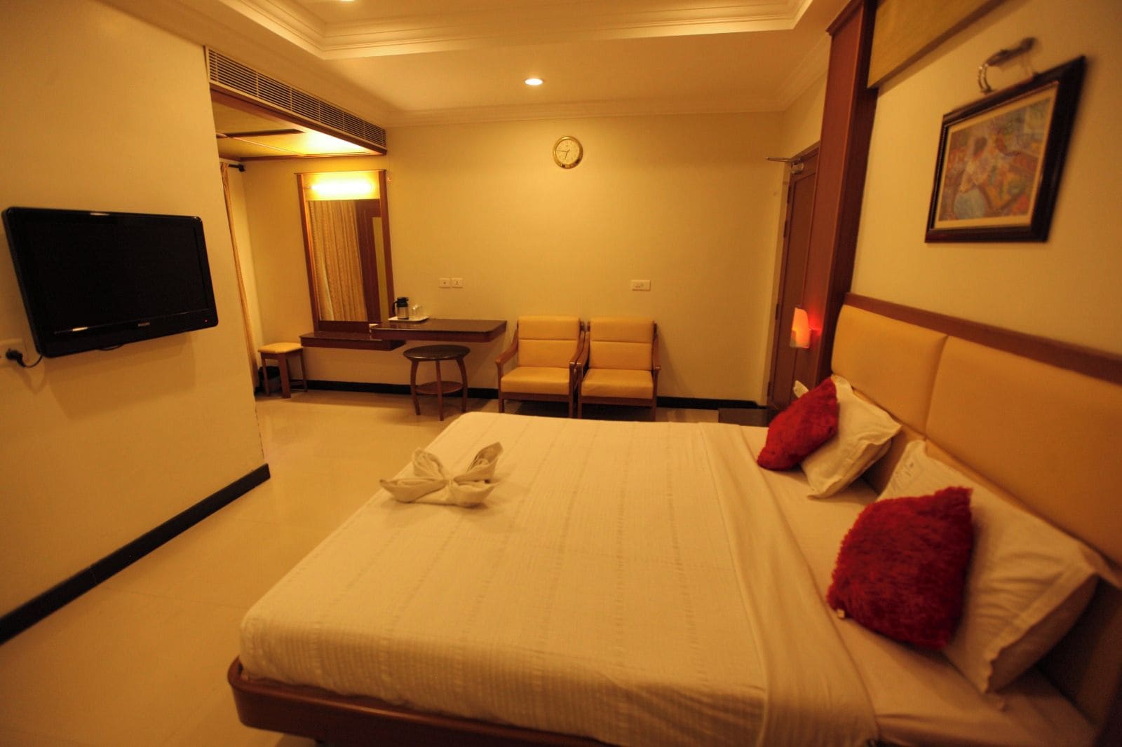 Quarto de hotel com cama, escrivaninha, cadeira, televisão e relógio na parede.