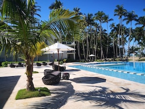 Área de piscina com espreguiçadeiras, guarda-sóis e palmeiras.