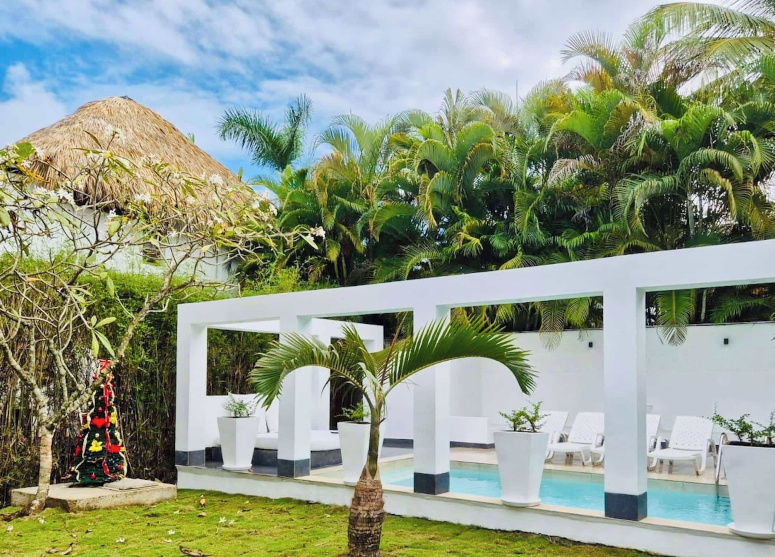 Uma área de piscina com espreguiçadeiras, uma estrutura com telhado de palha e vegetação tropical exuberante.