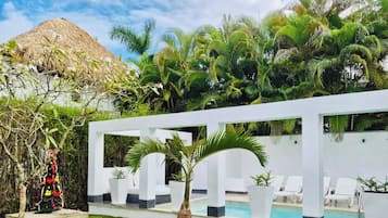 Uma área de piscina com espreguiçadeiras, uma estrutura com telhado de palha e vegetação tropical exuberante.