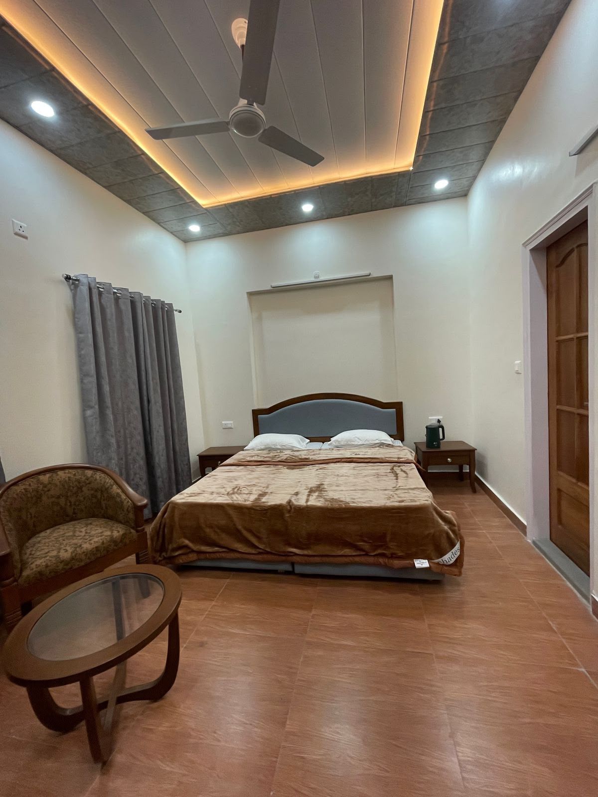 Quarto com cama, cadeira, mesa, ventilador de teto e janela com cortinas.
