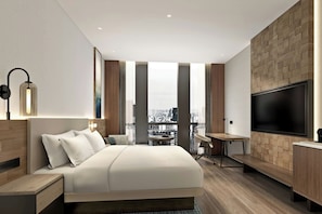 Quarto de hotel moderno com uma cama grande, escrivaninha, televisão e vista para a cidade.