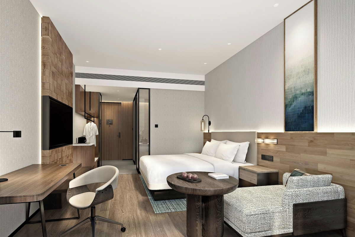 Quarto de hotel moderno com cama, mesa de cabeceira, cadeira e uma pequena mesa redonda.