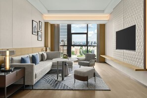 Sala de estar moderna com sofá, mesa de centro e poltrona, televisão de tela plana fixada na parede e vista para a cidade através de janelas amplas.