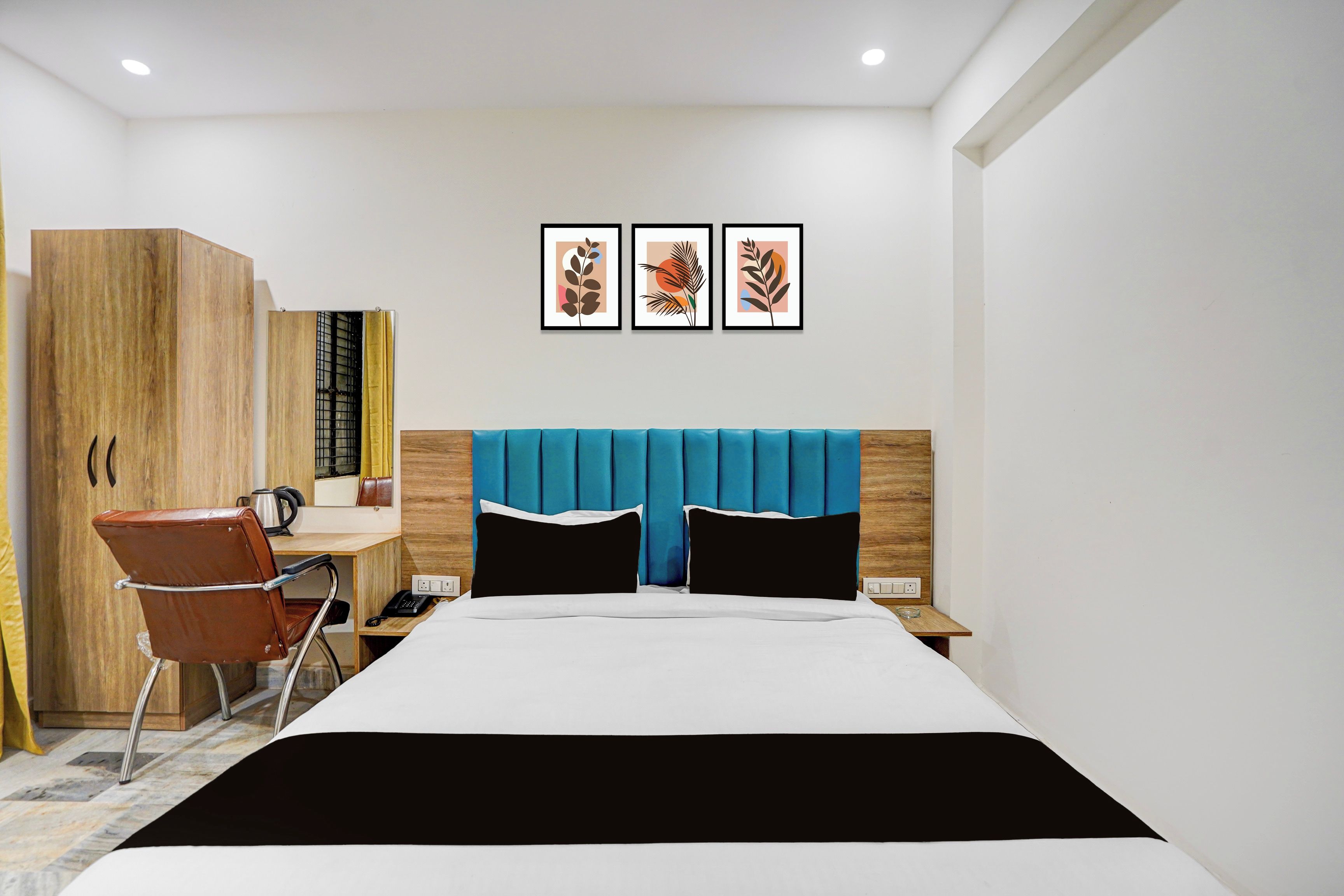 Quarto de hotel moderno com uma cama, escrivaninha, cadeira e guarda-roupa. Há três obras de arte emolduradas na parede.