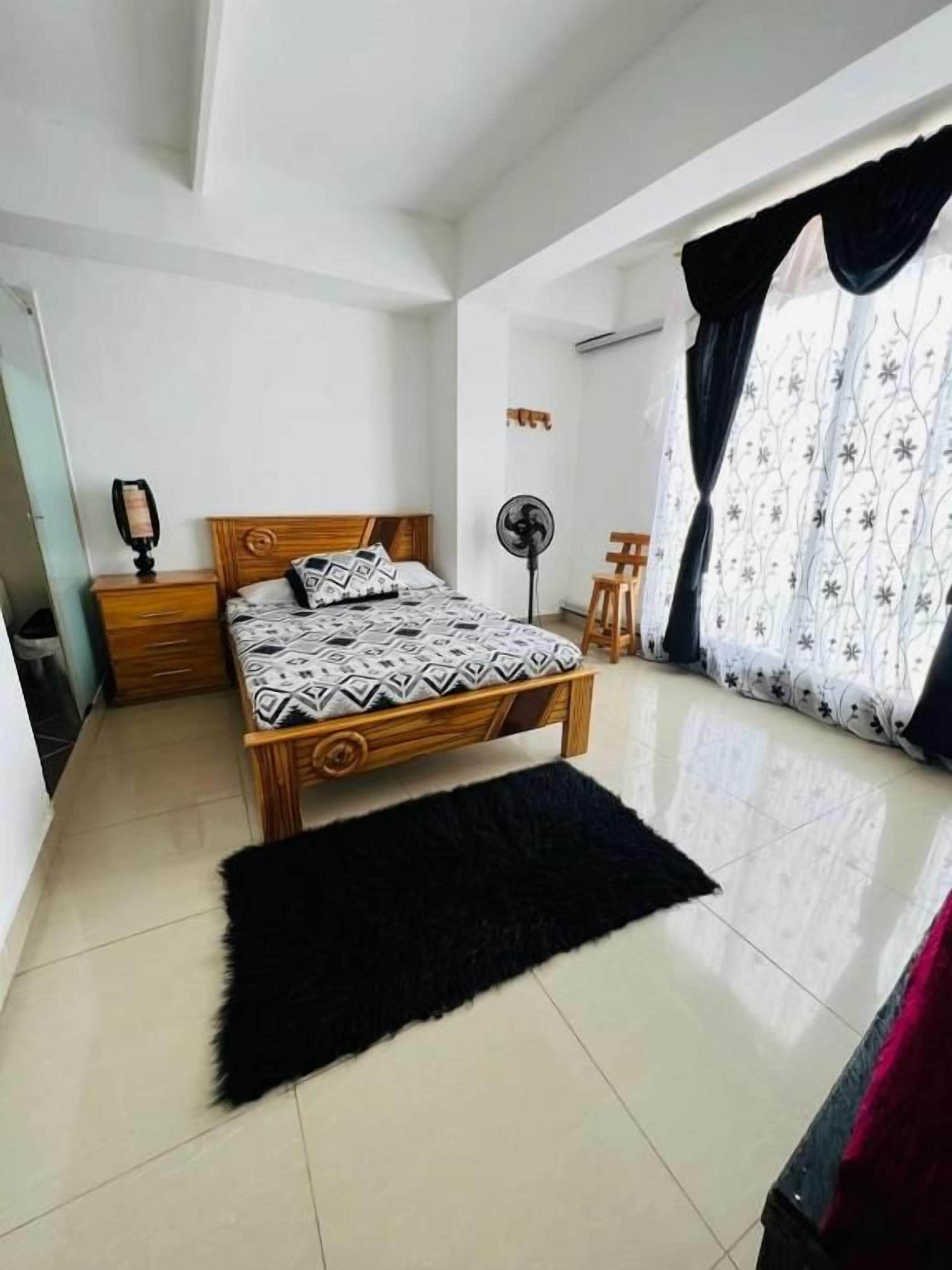 Quarto com cama, ventilador, tapete preto e janela com cortinas.