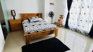 Quarto com cama, ventilador, tapete preto e janela com cortinas.